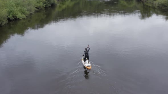 Aerial follows stand up paddleboarder paddling on small rural river ...