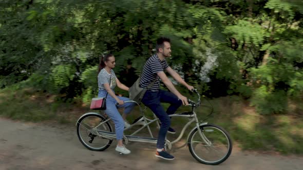 A young couple rides a tandem two-seater bike along the riverbank ...