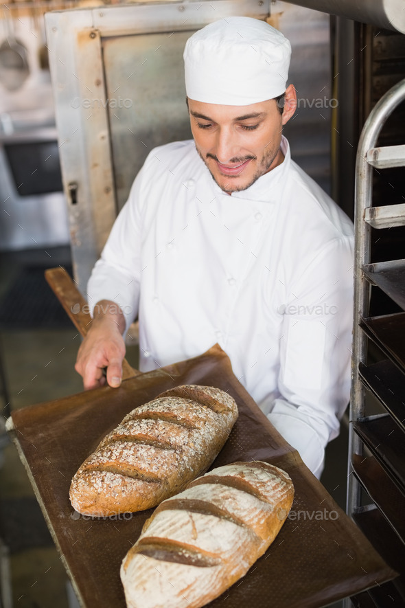 Happy baker holding tray of fresh bread in the kitchen of the bakery ...