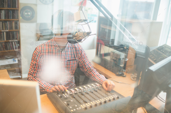 Radio host operating sound mixer in studio seen through glass Stock ...