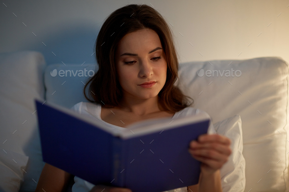 young woman reading book in bed at night home Stock Photo by dolgachov
