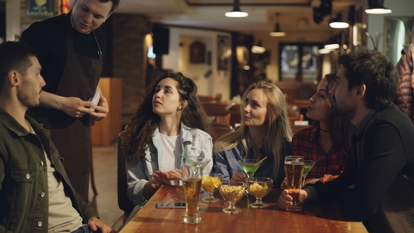 Handsome Male Waiter Is Coming To Group of Friends Sitting at Table and ...