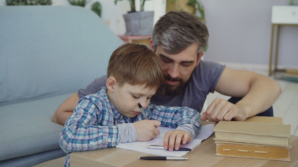 Cute Child Is Writing in Notebook with His Father Sitting Near the ...