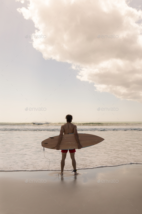 Rear view of shirtless young male surfer with surfboard standing on ...