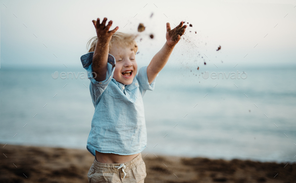 A small toddler boy standing on beach on summer holiday, throwing sand ...