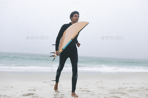 Front view of young mixed-race male surfer running on the beach with ...
