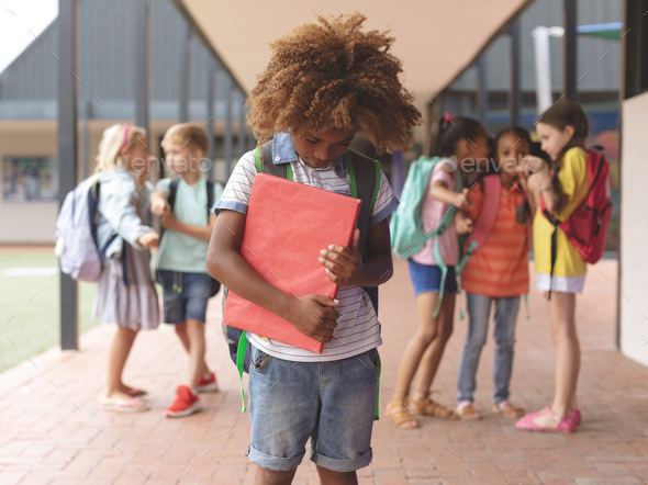 Sad schoolboy standing in corridor while being judged by his classmates ...