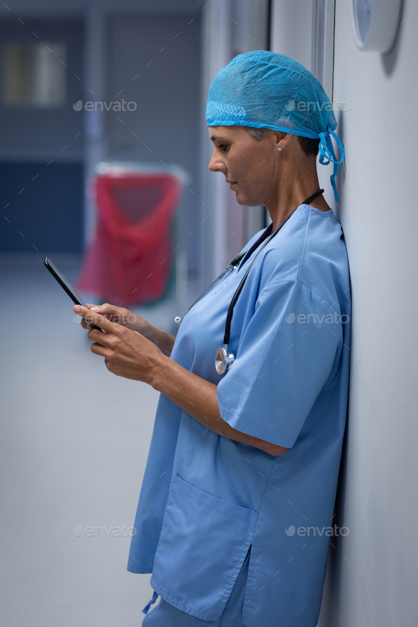 Female surgeon using mobile phone while leaning against a wall in ...