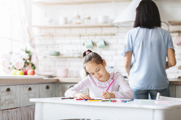 Schoolgirl doing homework in kitchen while her mother cooking food ...