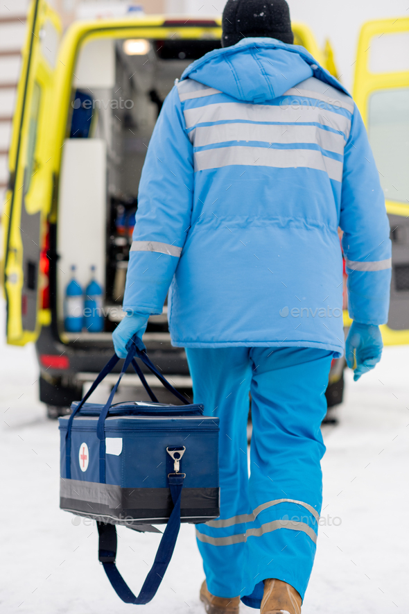 Rear view of paramedic in blue workwear and gloves carrying first aid ...
