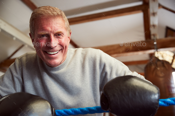 Smiling Senior Male Boxer In Gym Wearing Boxing Gloves Leaning On Ropes ...