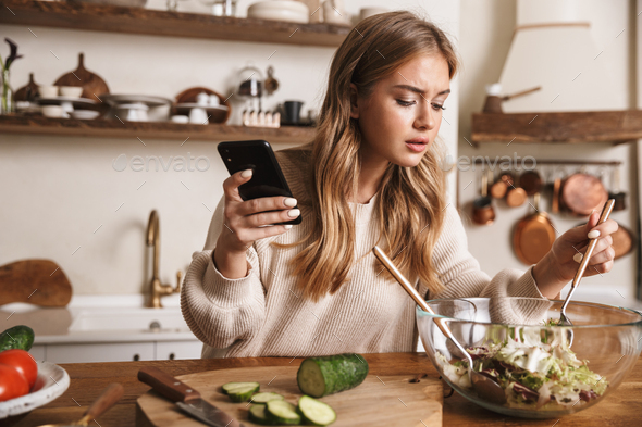 Image of thinking cute woman using cellphone while making lunch Stock ...