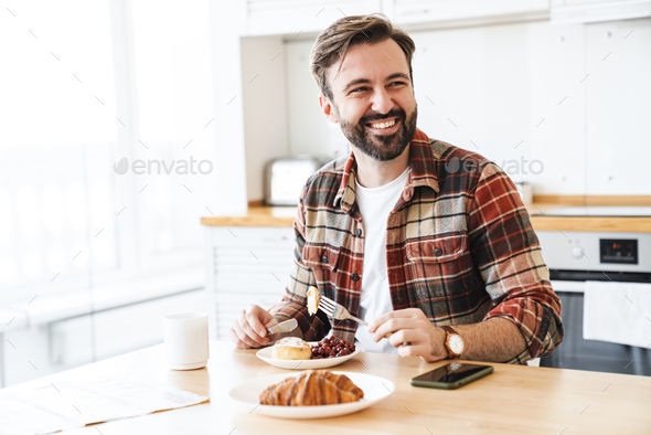Portrait of joyful man smiling and eating cake while having breakfast ...