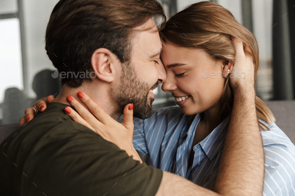 Photo of cheerful couple smiling and hugging while sitting on sofa ...