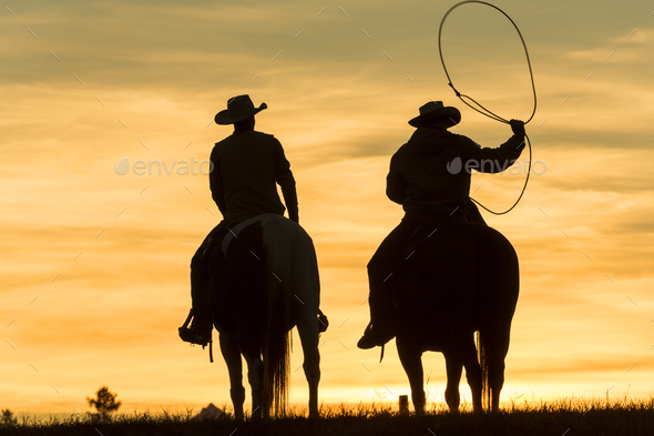 Two cowboys riding on horseback in a Prairie landscape at sunset, one ...