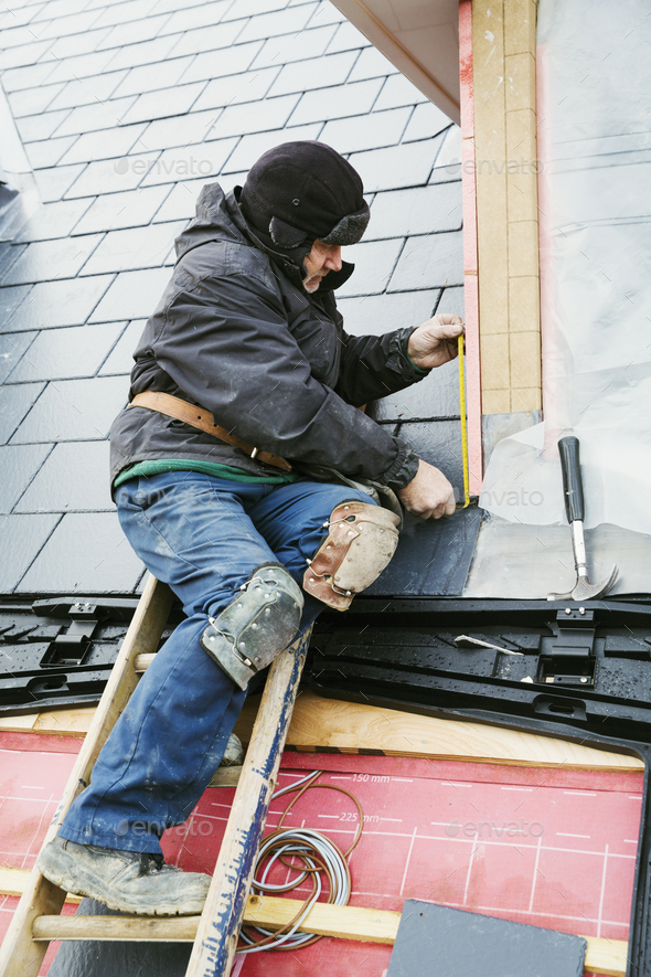 A man at the top of a ladder on a house roof, fixing tiles on a dormer ...