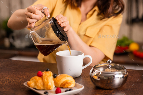 Young female sitting by table in the kitchen and pouring black tea into ...