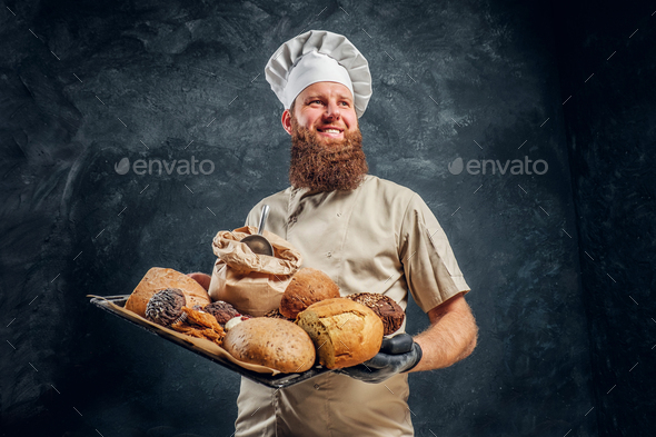 Bearded baker wearing a uniform standing next to a table with delicious ...