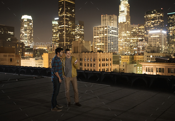 Two men on a rooftop overlooking Los Angeles at night, looking at a ...