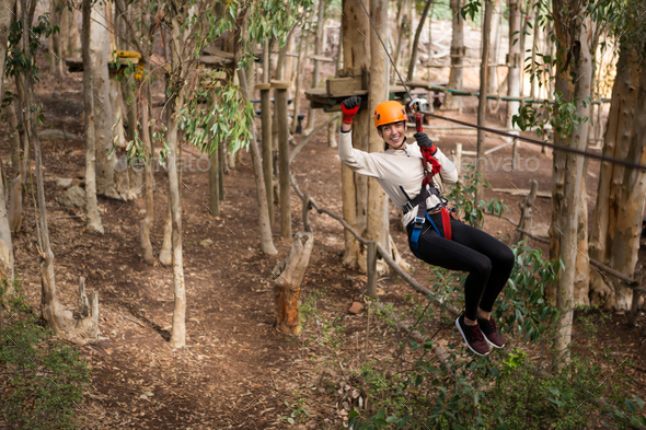 Happy woman wearing safety helmet riding on zip line in the forest ...