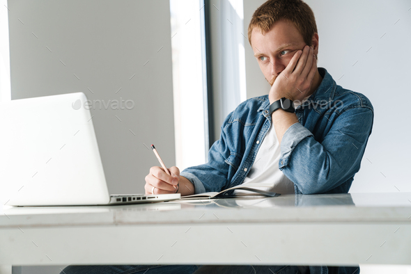 Photo of thinking man writing down notes while working with laptop ...