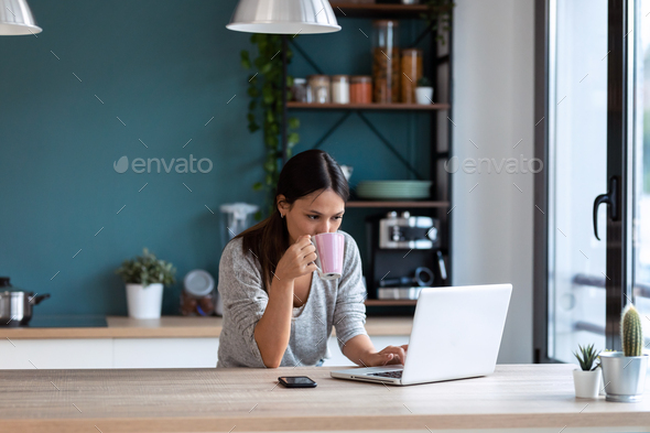 Pretty young woman drinking a cup of coffee while working with laptop ...