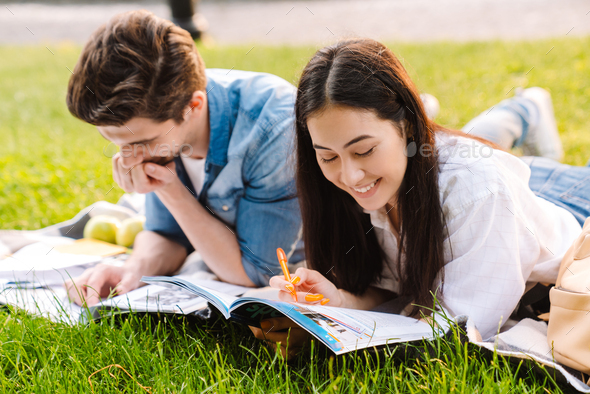 Image of joyful student couple doing homework while lying on grass ...