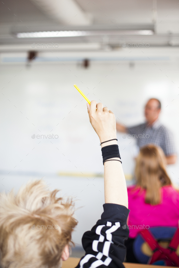 Rear view of schoolboy raising hand while teacher explaining in ...