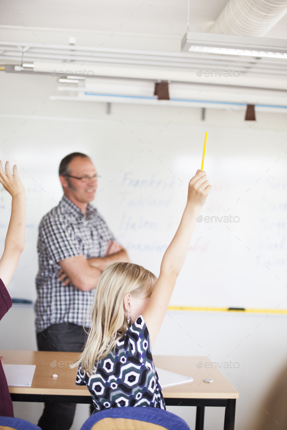 Schoolgirl raising hand while teacher explaining in classroom Stock ...