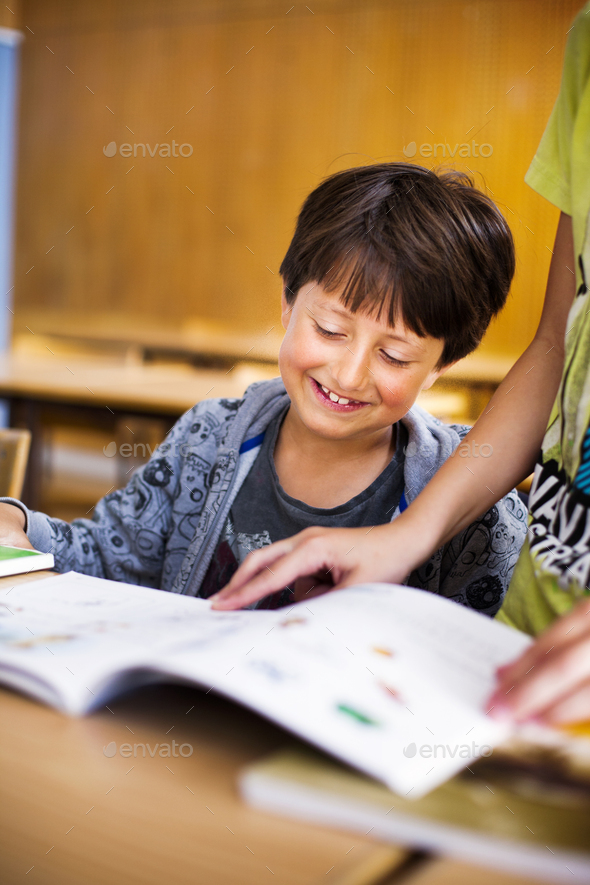 Smiling boy reading book while classmate standing at desk in classroom ...