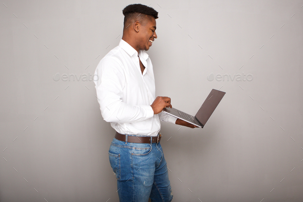 Profile of happy young black man holding laptop computer by gray wall ...
