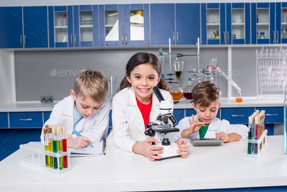 Three kids in chemical laboratory making notes and using microscope and ...