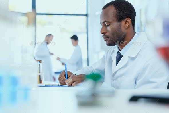 African american scientist in white coat taking notes while working in ...