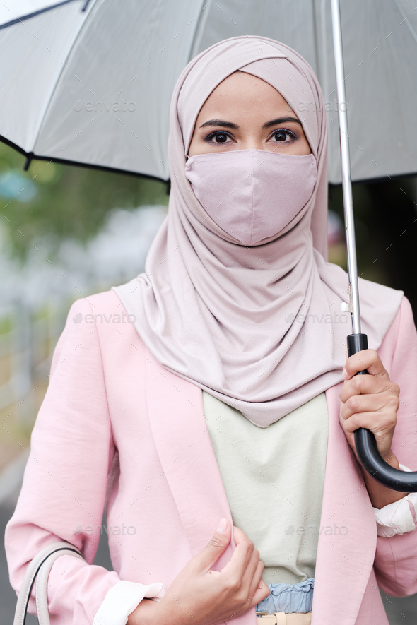Young Muslim woman in protective mask standing under umbrella on rainy ...