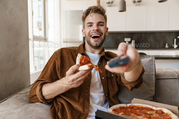Cheerful guy eating pizza while watching tv and using remote control ...
