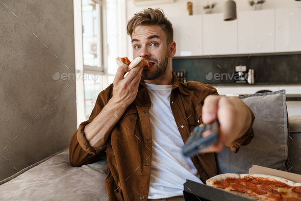 Excited guy eating pizza while watching tv and using remote control ...