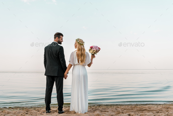 back view of wedding couple standing on beach with wedding bouquet and ...