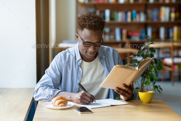 Funky black guy with textbook taking notes in copybook at cozy cafe ...