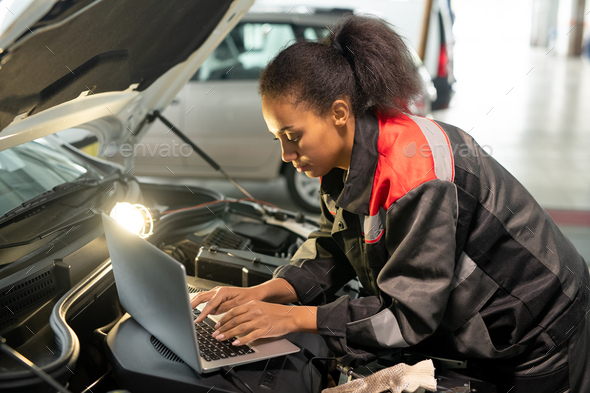 Female worker of car repair service using laptop over open engine ...