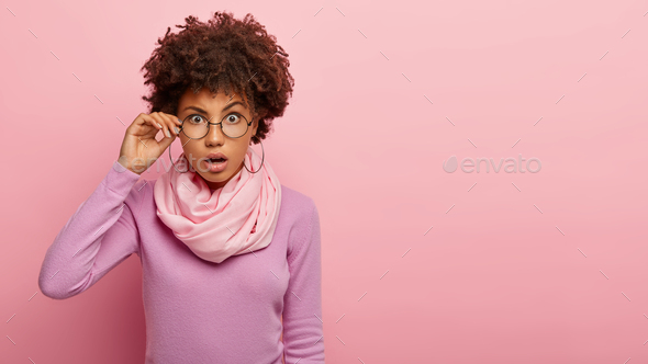 Young dazed woman with curly hair, keeps hand on frame of spectacles ...