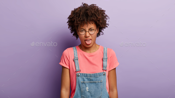 Photo of young curly woman feels disgust, sticks out tongue, sees ...