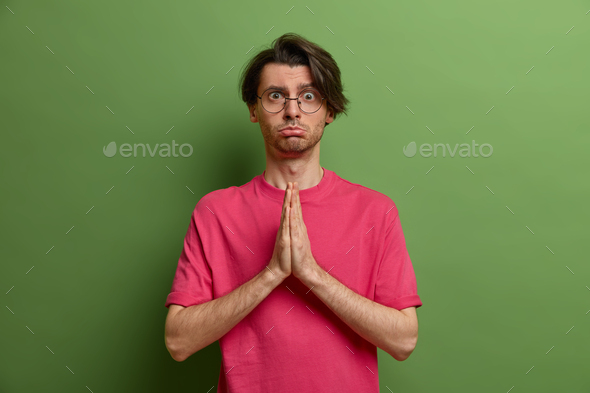 Sad adult man with imploring look, keeps palms pressed together, prays ...