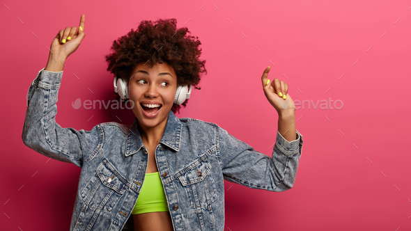 Indoor Shot Of Optimistic Dark Skinned Woman Dances And Keeps Arms Raised Moves With Rhythm Of
