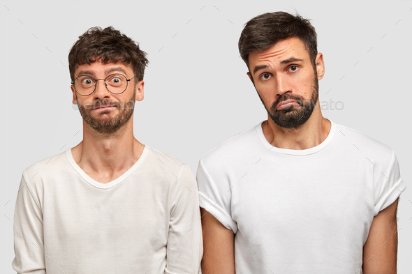 Puzzled sad two bearded young guys stand closely to each other, express ...