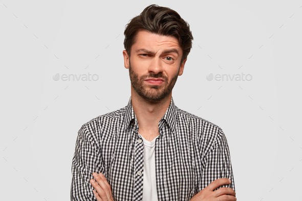 Stylish hipster male keeps arms folded, looks with hesitant expression ...