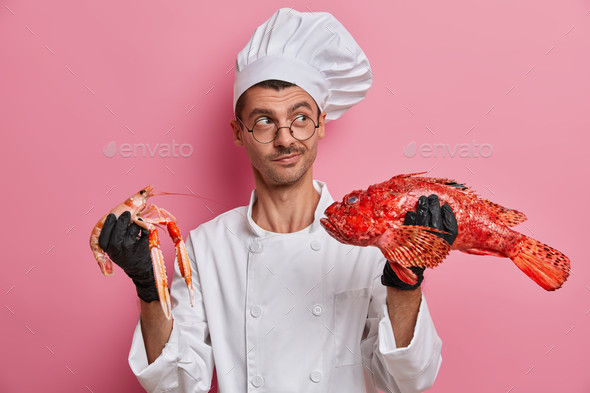 Serious pensive male chef in white uniform, holds crayfish and red sea bass, prepares for sreet ...
