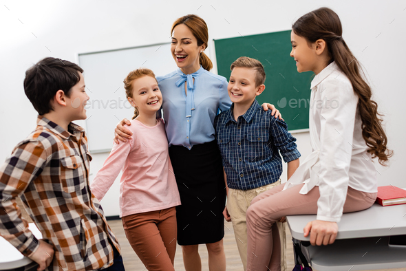 Laughing teacher embracing pupils in front of blackboard in classroom ...