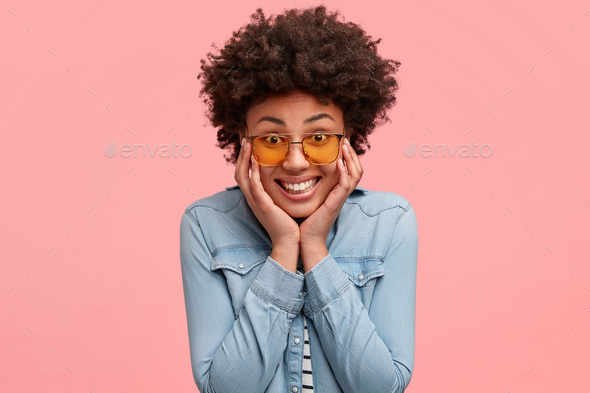 Delighted positive dark skinned beautiful female touches cheeks, smiles ...