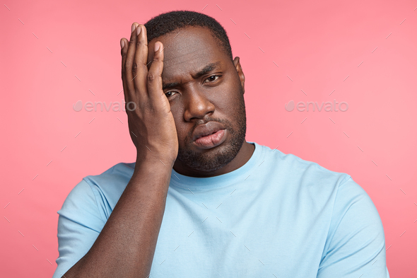Headshot of depressed gloomy disappointed black man rests hand on head, has headache after long ...