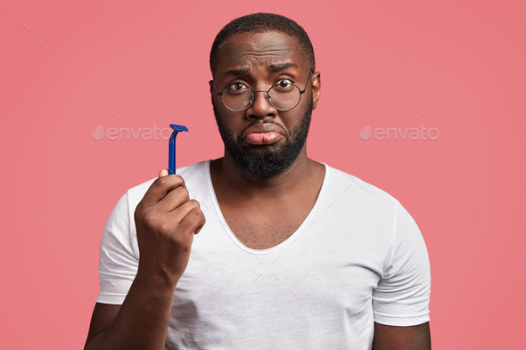 Unhappy dark skinned man holds razor, doesn`t want to shave his long ...